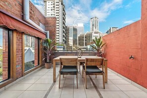 Um terraço no telhado com mesa e cadeiras de madeira, plantas em vasos e vista para a cidade.