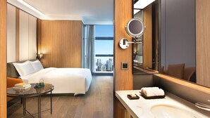 Quarto de hotel com uma cama, um espelho redondo, uma mesinha com livros, um banheiro com pia e vista para a cidade.