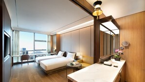 Quarto de hotel com duas camas, área de estar, banheiro com espelho e pia, e vista para a cidade.
