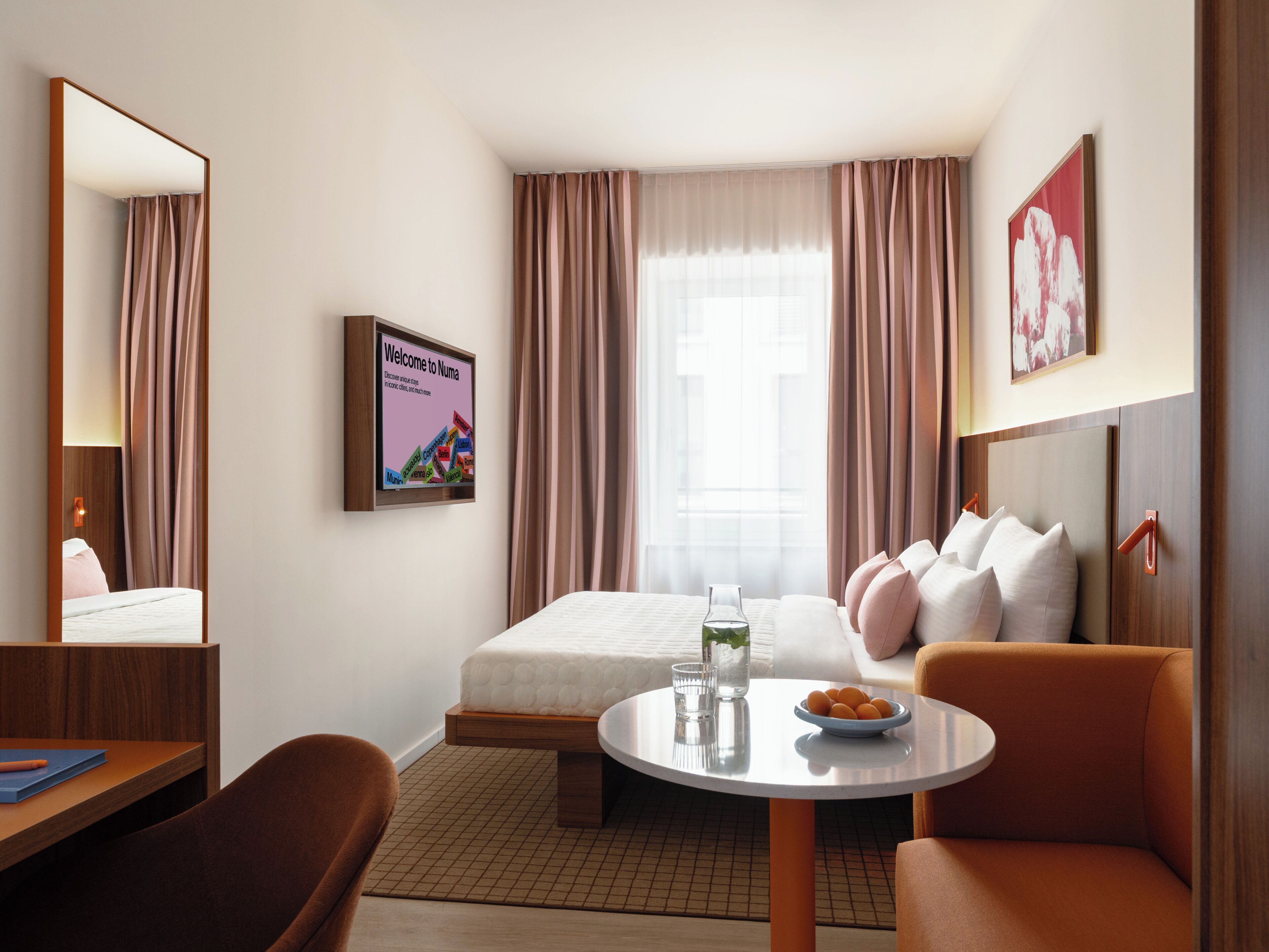 Quarto de hotel com cama, escrivaninha, cadeira, mesa redonda com um vaso e uma tigela de frutas, espelho, placa de boas-vindas e janela com cortinas.