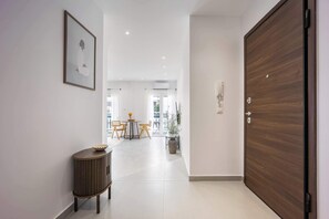 Um corredor com porta de madeira, quadro na parede, mesa redonda pequena com uma tigela e planta em vaso.