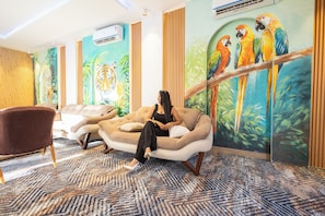 Um quarto com um grande mural de uma tigresa e papagaios, uma poltrona marrom, um sofá e um tapete estampado.