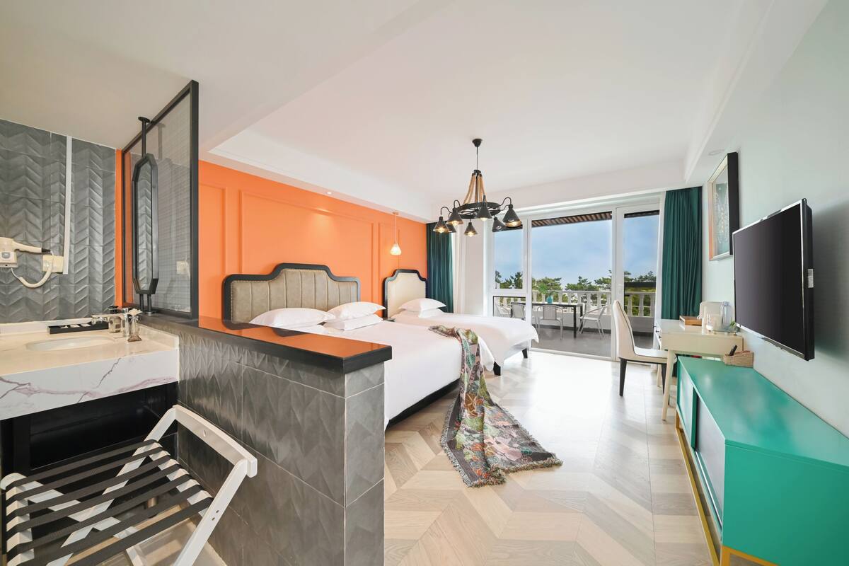 Quarto de hotel com cama, escrivaninha, televisão, banheiro com chuveiro e varanda com vista.