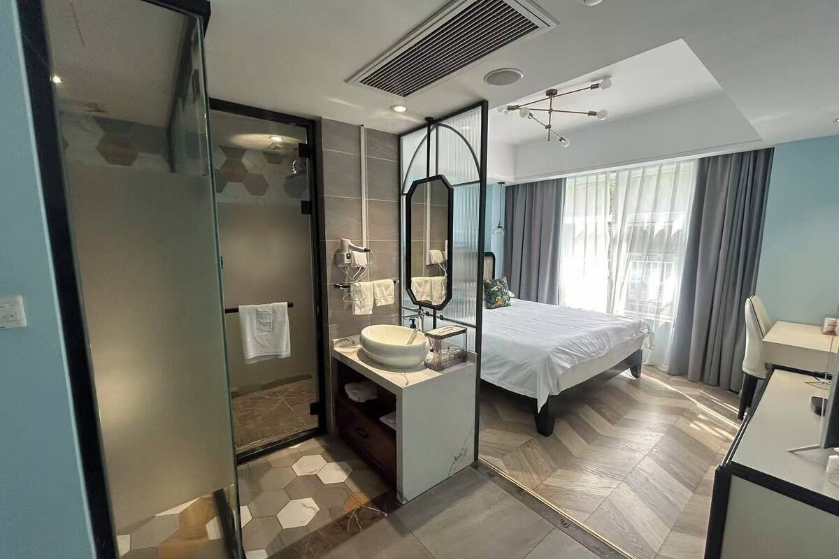 Quarto de hotel moderno com chuveiro em cabine de vidro, pia redonda, cama grande e escrivaninha com cadeira.