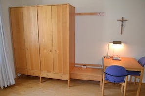 Um quarto com um guarda-roupa de madeira, uma escrivaninha com cadeira, uma cruz na parede e um abajur.