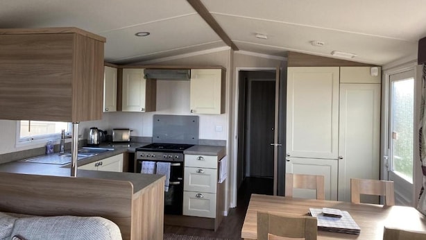 Cozinha compacta de RV com fogão, pia e armários, área de jantar com mesa e cadeiras, e uma porta que dá acesso a outro cômodo.