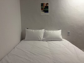 Quarto simples com uma cama, duas almofadas, um interruptor de luz fixado na parede e um cartaz.