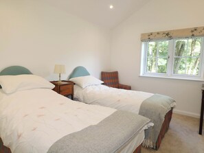 Quarto com duas camas, uma mesa de cabeceira com luminária, janela com vista para vegetação e uma cadeira.