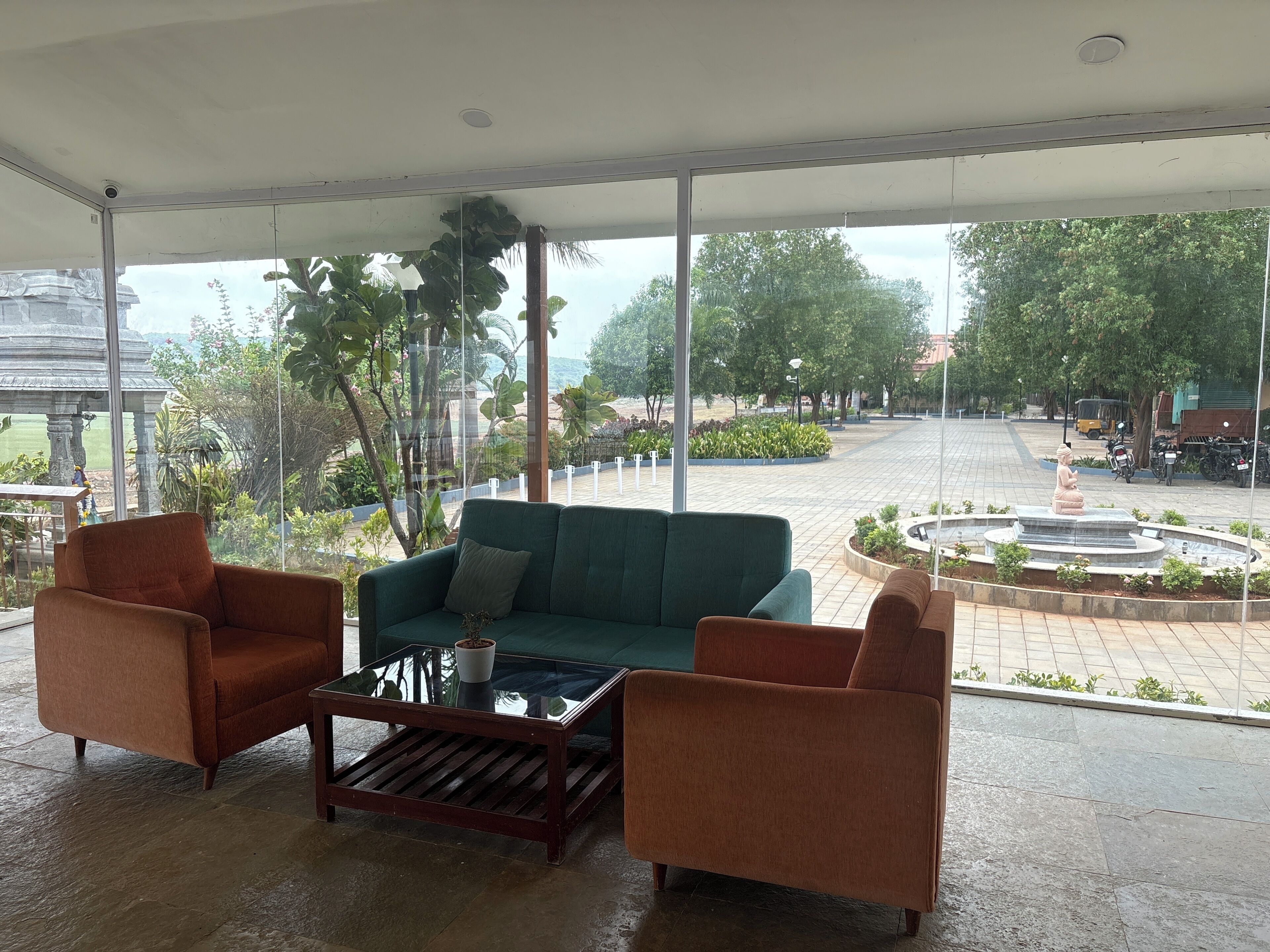 Uma área de estar coberta com sofá e poltronas, uma mesa de centro e vista para um pátio com uma estátua e vegetação.
