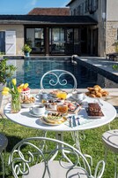 Disposição para café da manhã à beira da piscina, com mesa, cadeiras e diversas opções de comida e bebidas.