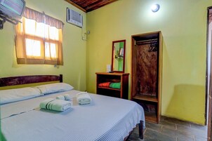 Quarto de hotel com cama, espelho, prateleira com toalhas e guarda-roupa de madeira.