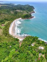 Vista aérea de uma área costeira com praia, vegetação densa e alguns edifícios.