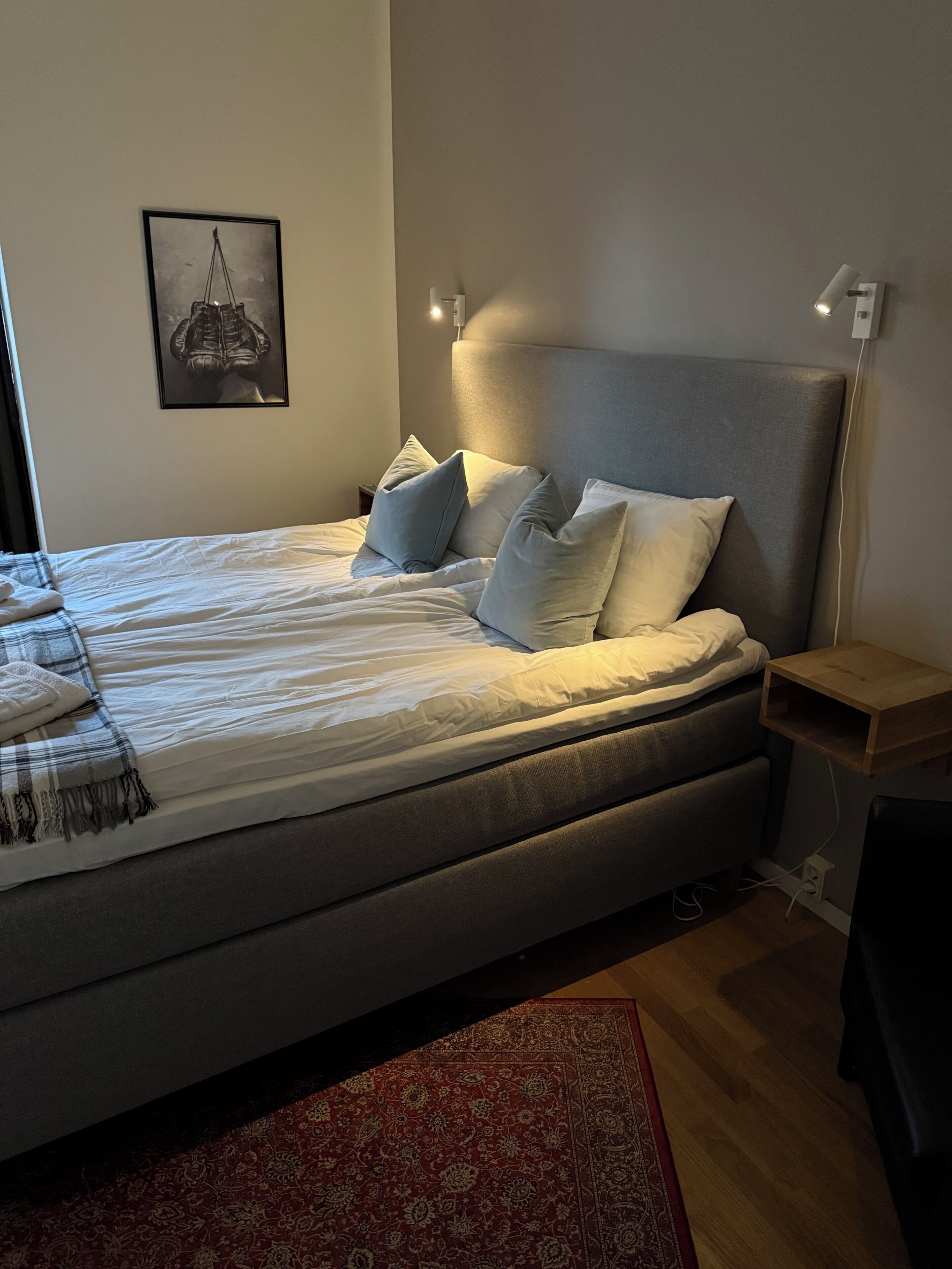 Uma cama bem arrumada com almofadas e um cobertor, uma mesa de cabeceira com abajur, um quadro na parede e um tapete estampado no chão.