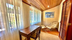 Um quarto com teto de madeira, uma mesa de jantar com cadeiras, uma poltrona estofada, um quadro na parede e uma porta com persianas de madeira.
