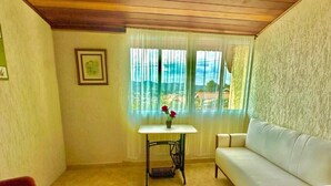 Um quarto com sofá branco, uma mesinha com uma máquina de costura, uma janela com cortinas e vista para prédios.