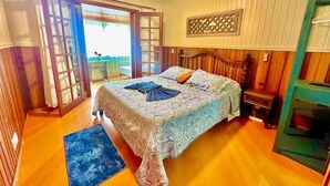 Quarto com cama, cabeceira de madeira, mesa de cabeceira, janela com cortinas e vista para o exterior.