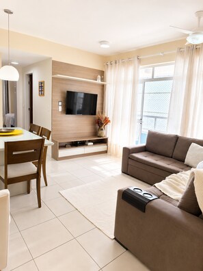 Uma sala de estar moderna com uma TV de tela plana, um sofá, uma mesa de jantar com cadeiras e uma janela grande com cortinas.