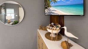 Quarto moderno com TV de tela plana exibindo uma cena de praia, um espelho redondo, uma tigela decorativa com conchas e um armário de madeira.