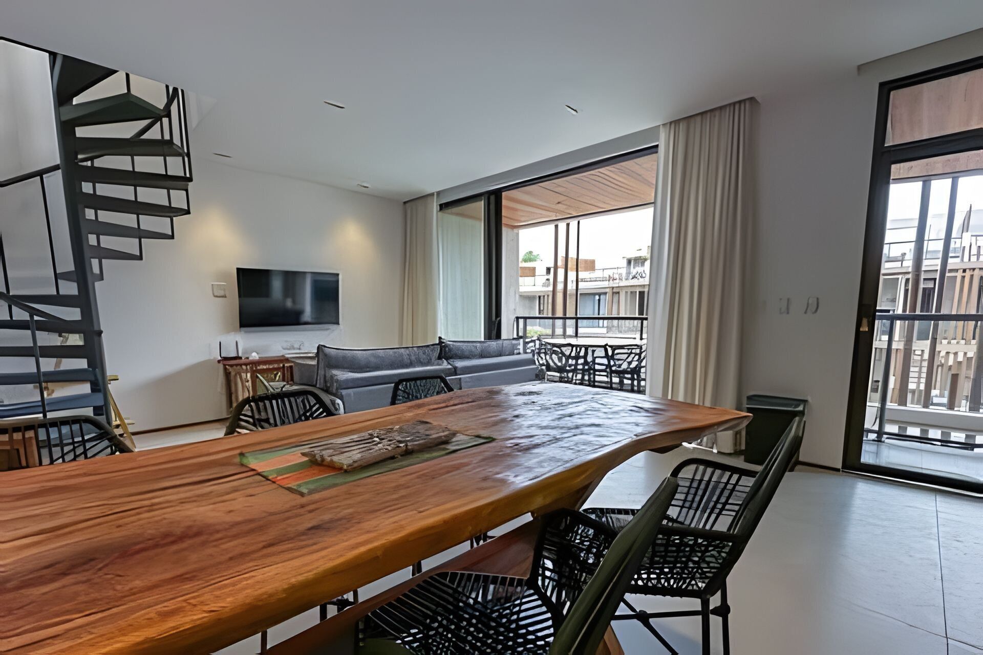 Uma sala de estar moderna com uma mesa de jantar de madeira, cadeiras pretas, um sofá cinza, uma TV e uma escada.
