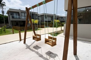 Um parquinho com baloiço numa área externa, com edifícios modernos ao fundo.