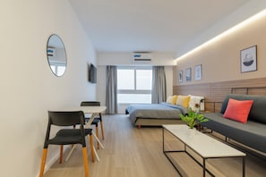 Quarto de hotel moderno com cama, sofá, mesa de jantar com cadeiras, espelho e planta em vaso.