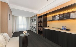 Quarto de hotel com sofá, uma pequena mesa redonda com um vaso, área de lavanderia com máquinas de lavar e secar, e uma mesa de recepção com painéis informativos.