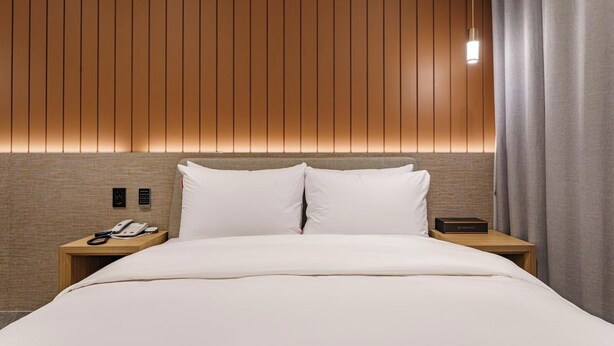 Quarto de hotel com cabeceira de madeira, cama com roupa branca, mesa de cabeceira com telefone e abajur, e cortina no lado direito.