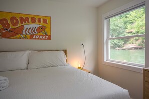 Quarto com cama, mesa de cabeceira com luminária, janela com vista para área verde e um objeto decorativo na parede.