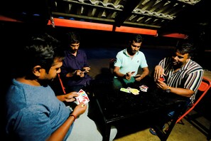 Quatro pessoas jogando cartas numa mesa num ambiente com pouca luz, com uma mesa preta e cadeiras vermelhas.