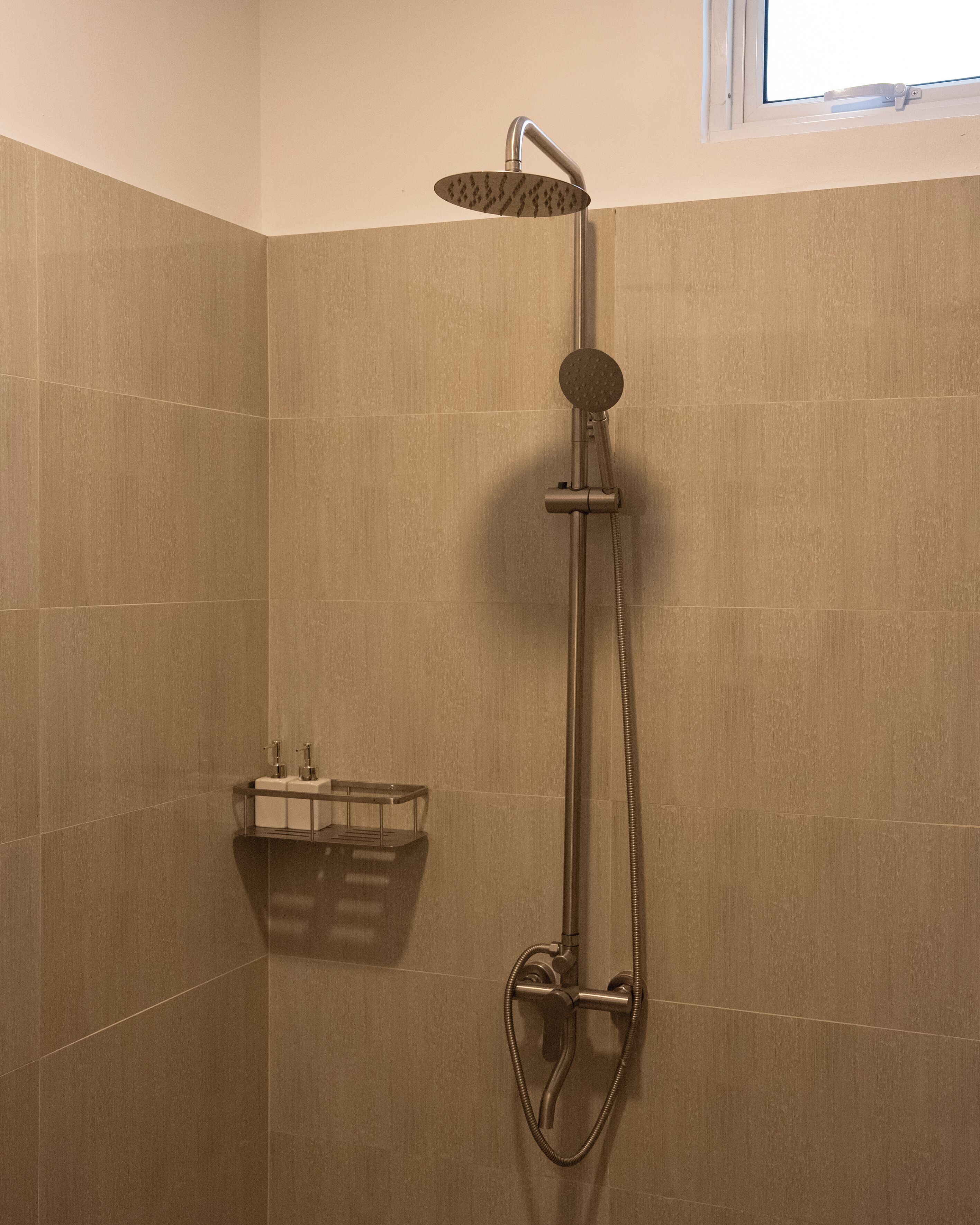 Um chuveiro com chuveiro manual e ducha de teto, um nicho pequeno com produtos de higiene e uma janela acima do chuveiro.