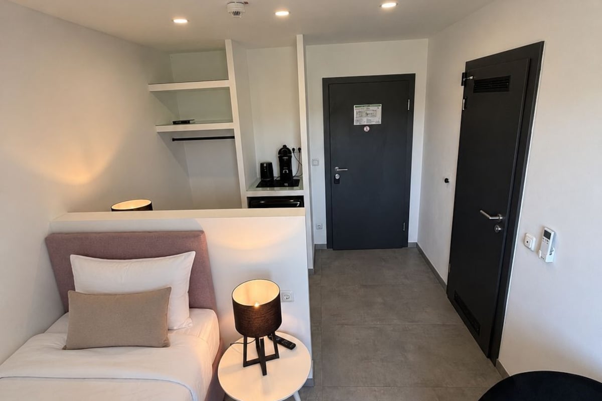 Quarto de hotel moderno com uma cama, uma mesinha com abajur, uma cadeira preta, uma porta escura e uma parede branca com prateleira.