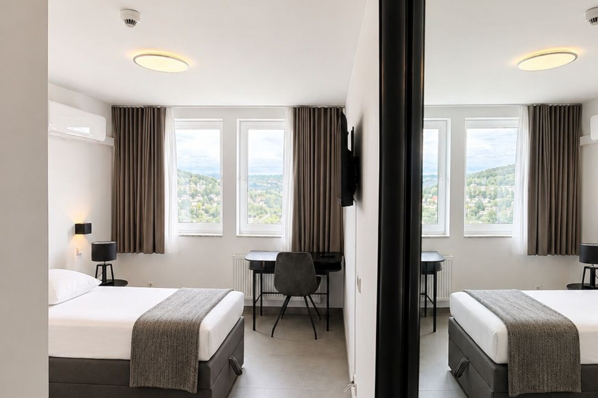 Quarto de hotel com duas camas, uma escrivaninha e uma cadeira. Janelas com cortinas, uma TV de tela plana fixada na parede e um ar condicionado na parede.
