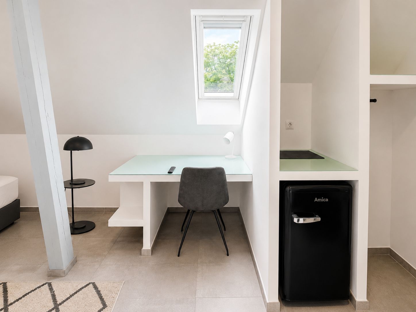 Quarto compacto com escrivaninha, cadeira, luminária e um pequeno eletrodoméstico preto, possivelmente uma geladeira.