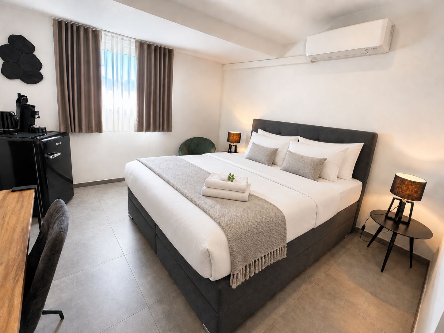 Quarto de hotel moderno com uma cama grande, uma escrivaninha, uma cadeira e uma mesinha de cabeceira com abajur.