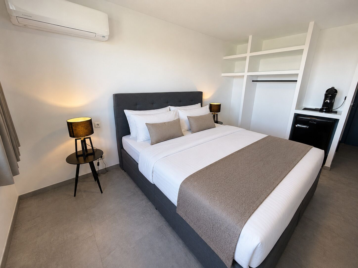 Quarto de hotel moderno com uma cama grande, luminárias de cabeceira, escrivaninha embutida e mini-geladeira.