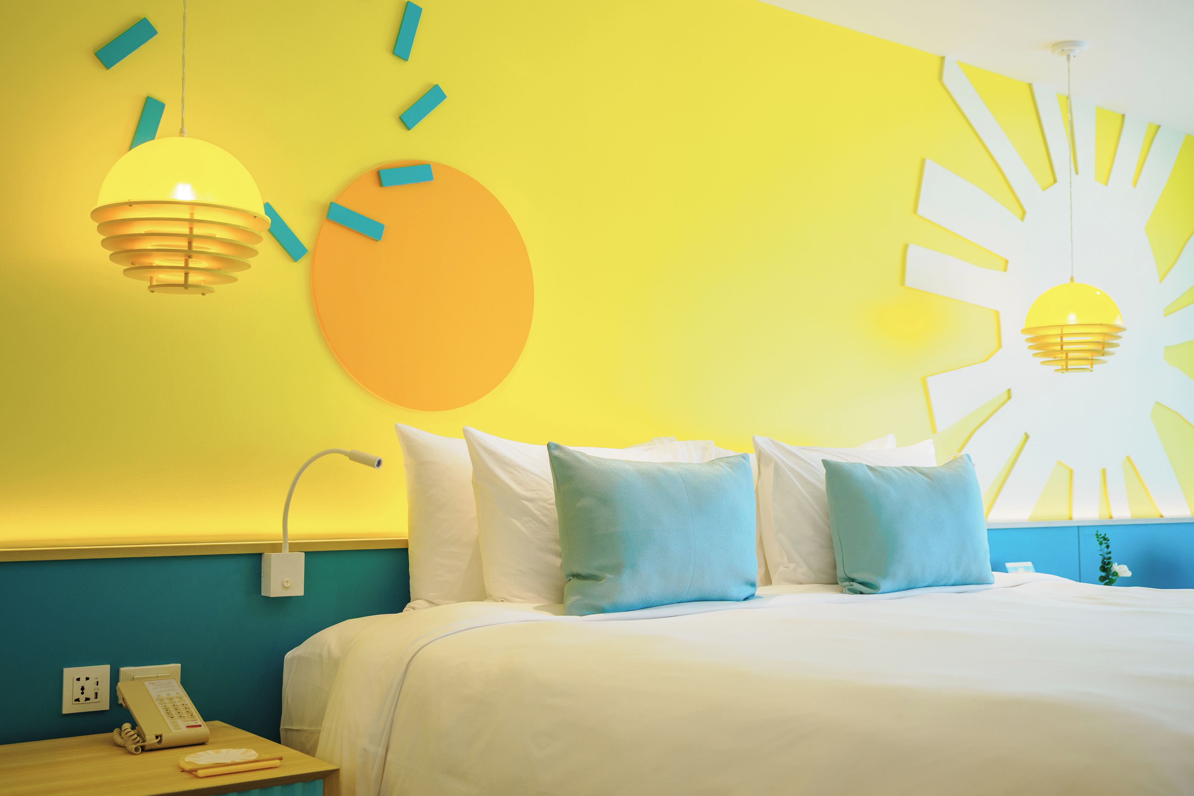 Quarto de hotel com cama, mesa de cabeceira e decoração na parede, incluindo um desenho de sol e luminárias pendentes.