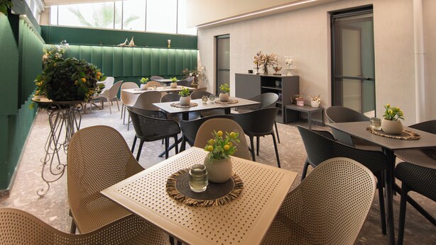 Um restaurante com várias mesas e cadeiras, uma área de bancos acolchoados verdes e plantas em vasos.