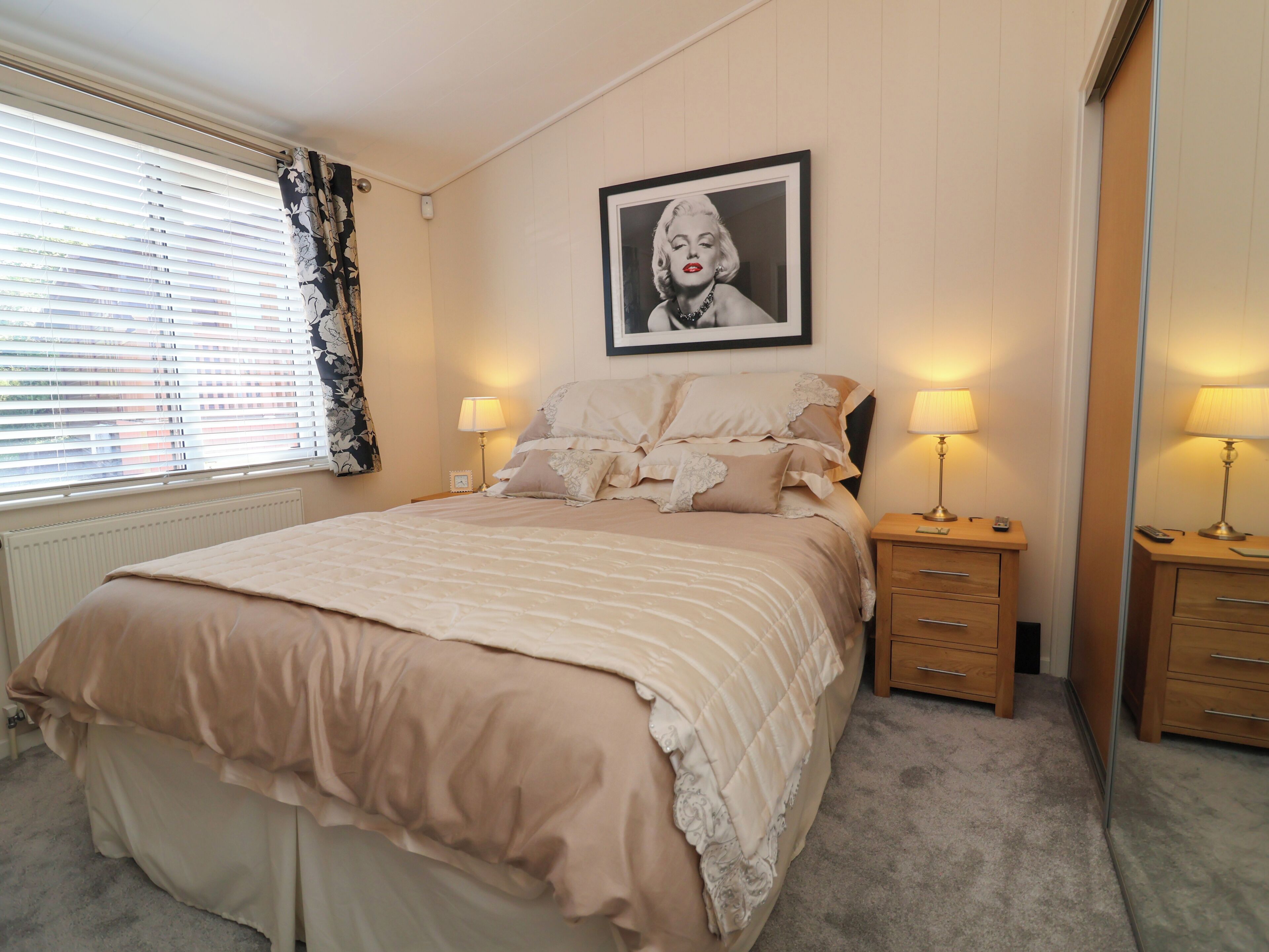 Quarto com cama, duas mesas de cabeceira, abajur, quadro na parede e janela com persianas.