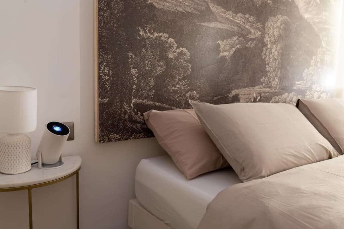 Quarto com cama, almofadas, abajur e dispositivo fixado na parede.