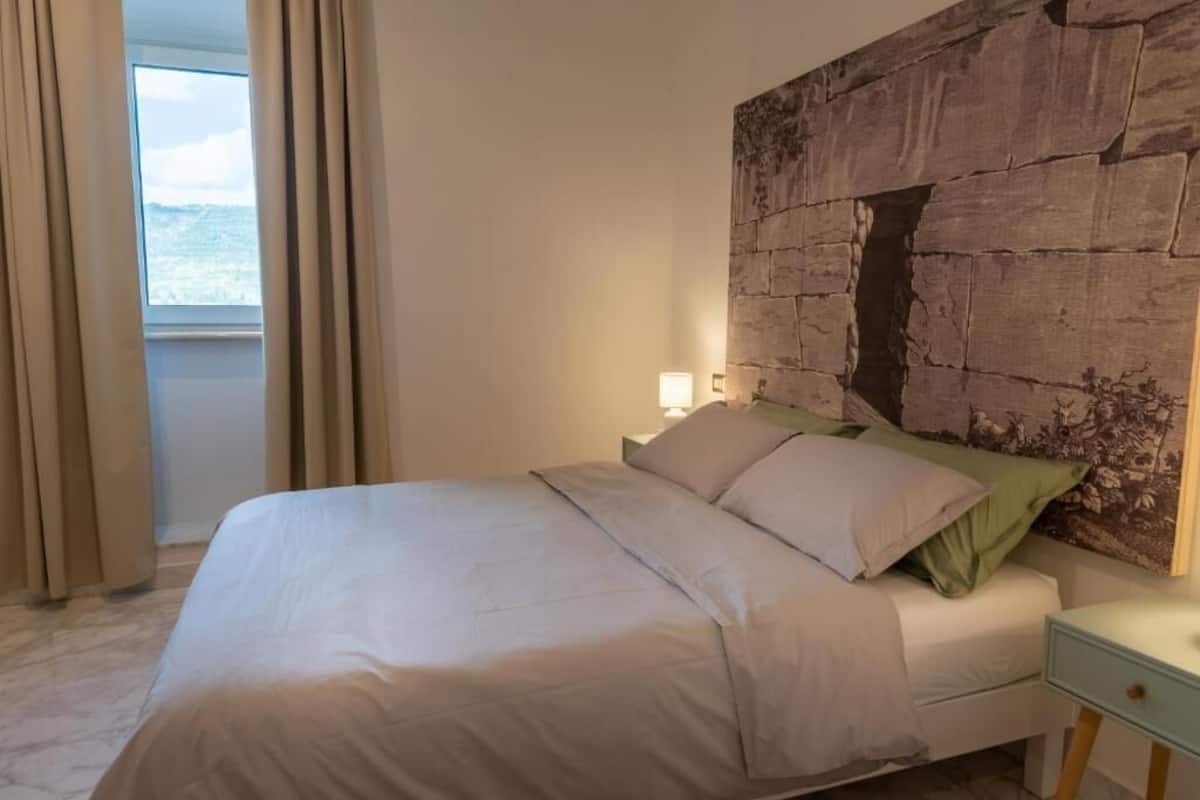 Quarto com cama, mesa de cabeceira, parede com textura e vista pela janela.