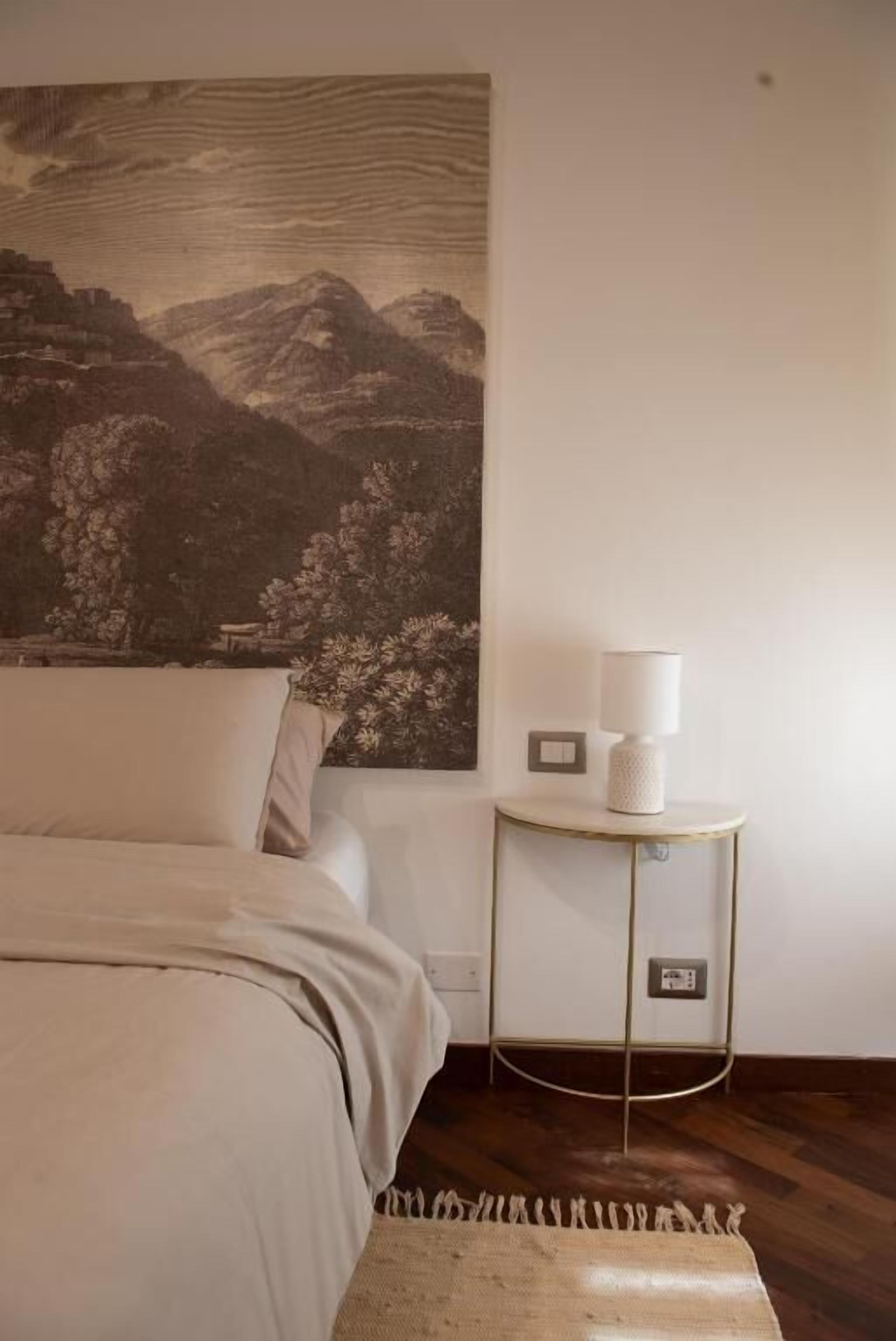 Quarto com cama, mesa de cabeceira com abajur, tomada na parede e uma grande pintura de paisagem.