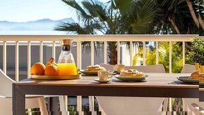 Mesa posta com itens de café da manhã, um copo de suco de laranja e uma garrafa, com vista para palmeiras e montanhas ao fundo.