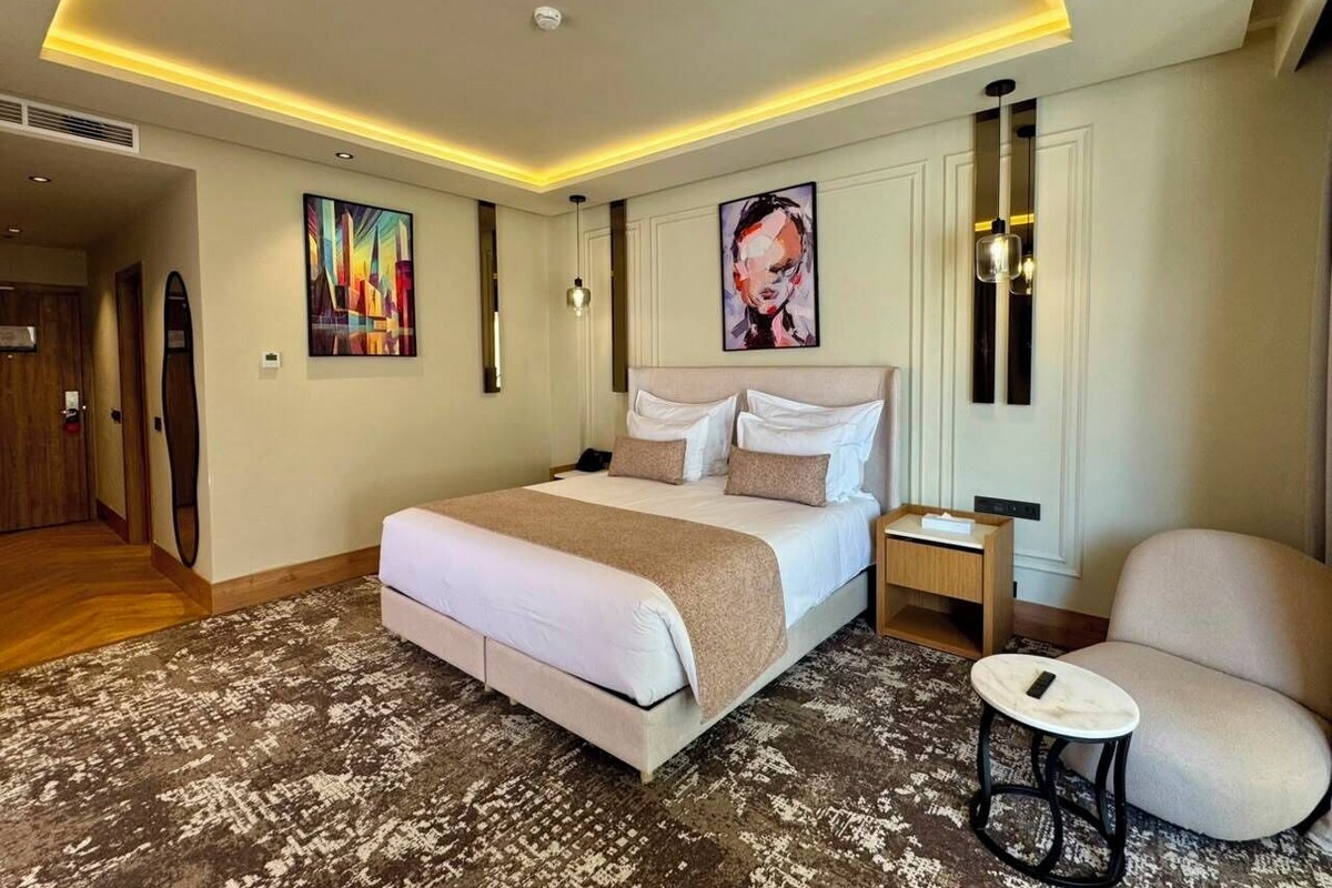 Quarto de hotel com uma cama grande, mesas de cabeceira, uma cadeira e obras de arte nas paredes.