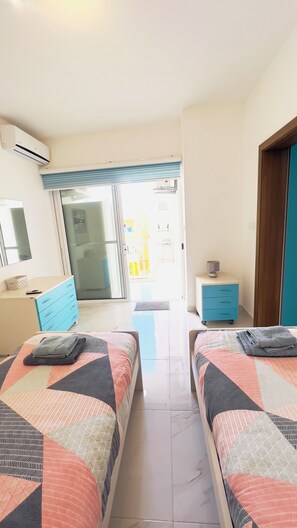 Quarto com duas camas, um guarda-roupa, mesa de cabeceira, espelho e porta de vidro deslizante para uma varanda.
