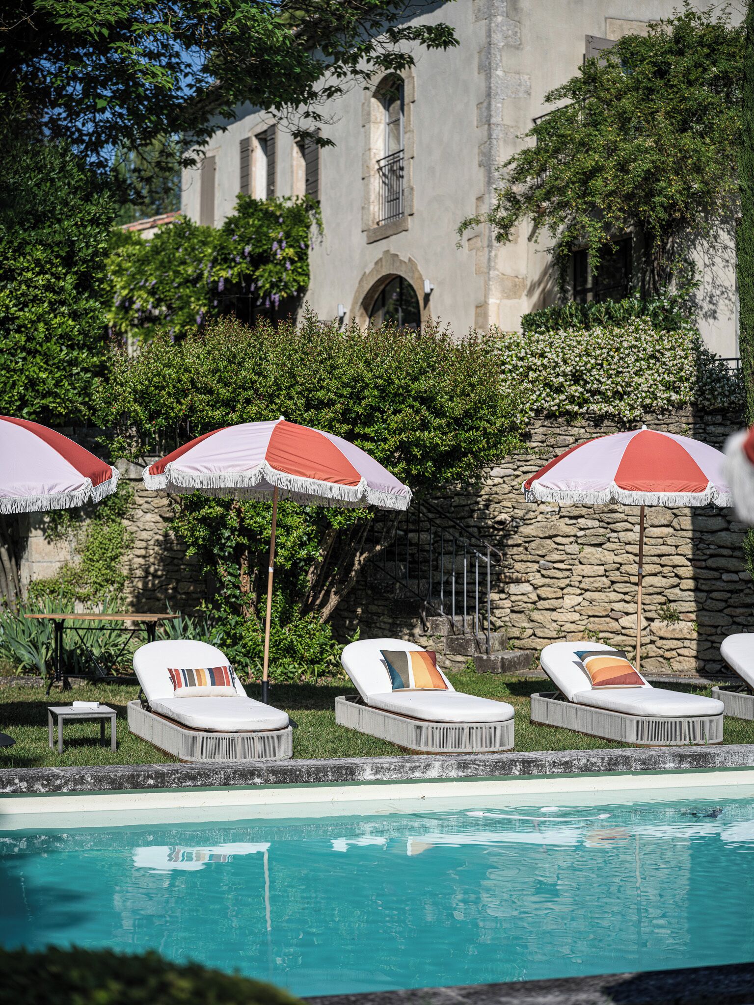 Uma área à beira da piscina com espreguiçadeiras e guarda-sóis, um muro de pedra e um edifício com janelas em arco ao fundo.