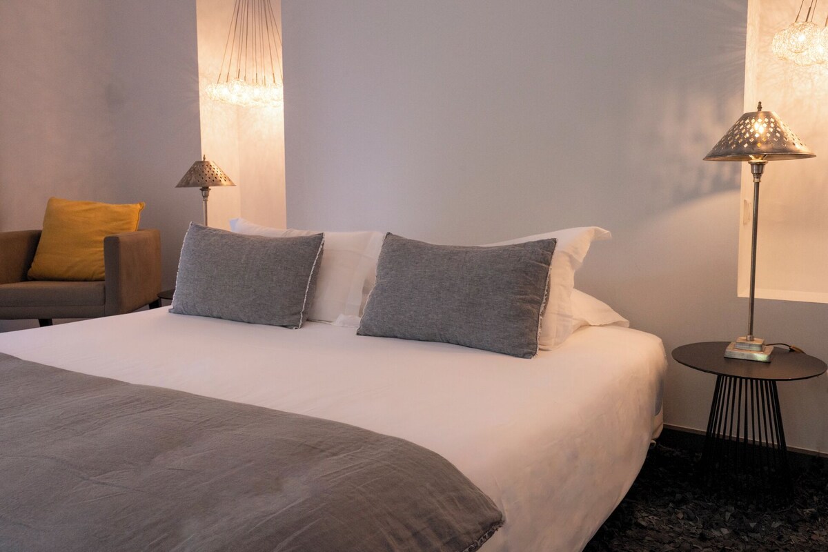 Quarto de hotel com uma cama grande, duas almofadas, mesa de cabeceira com abajur, cadeira com almofada e luminária na parede.