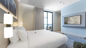 Quarto de hotel com cama, televisão, janela com cortinas e vista para o mar.