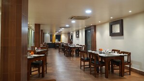 Um corredor extenso com várias mesas e cadeiras para refeições, painéis de madeira nas paredes e luminárias embutidas no teto.
