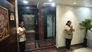 Duas pessoas em um saguão, com um elevador, um vaso de planta e um quadro na parede.