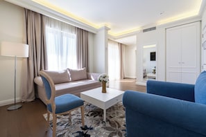 Quarto de hotel moderno com sofá, poltrona azul, mesa de centro branca e um guarda-roupa grande.